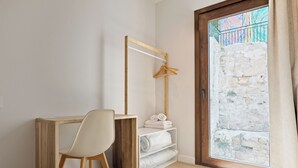 Quarto com uma secretária de madeira, uma cadeira branca e um guarda-roupa de madeira. Há uma janela com vista para uma parede de pedra e um portão colorido.