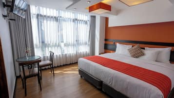 Quarto de hotel com uma cama grande, uma escrivaninha com cadeira, uma televisão na parede e uma janela com cortinas.