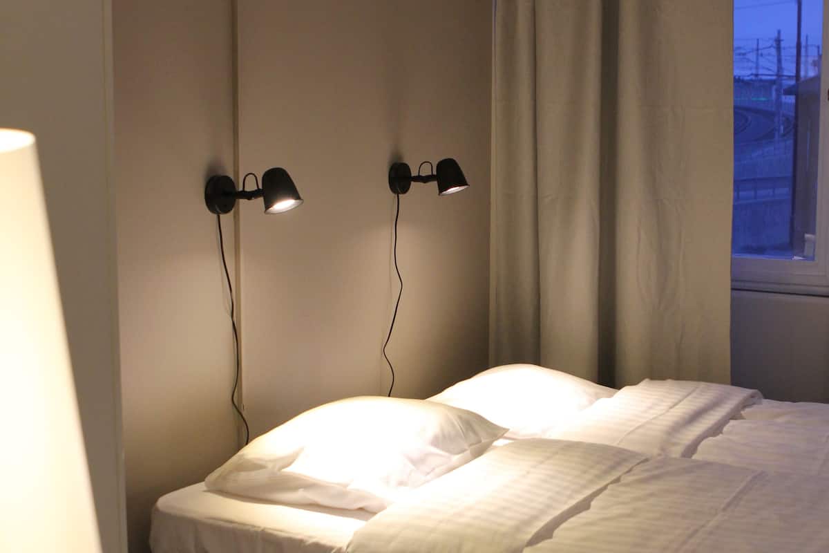 Quarto com cama, duas luminárias na parede, janela com cortinas e porta.