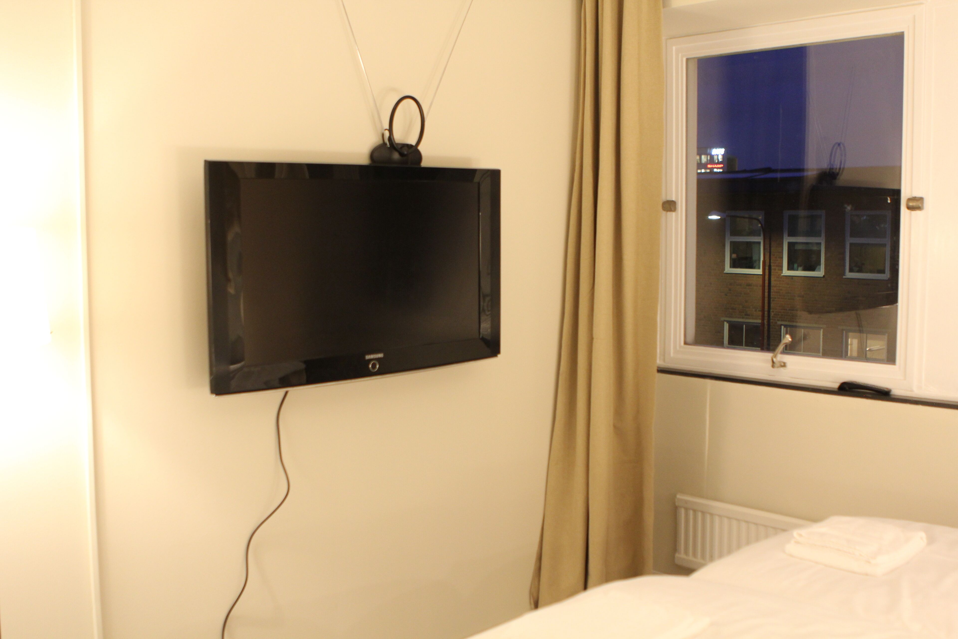 Quarto com televisão montada, janela com cortinas e cama com toalha dobrada.