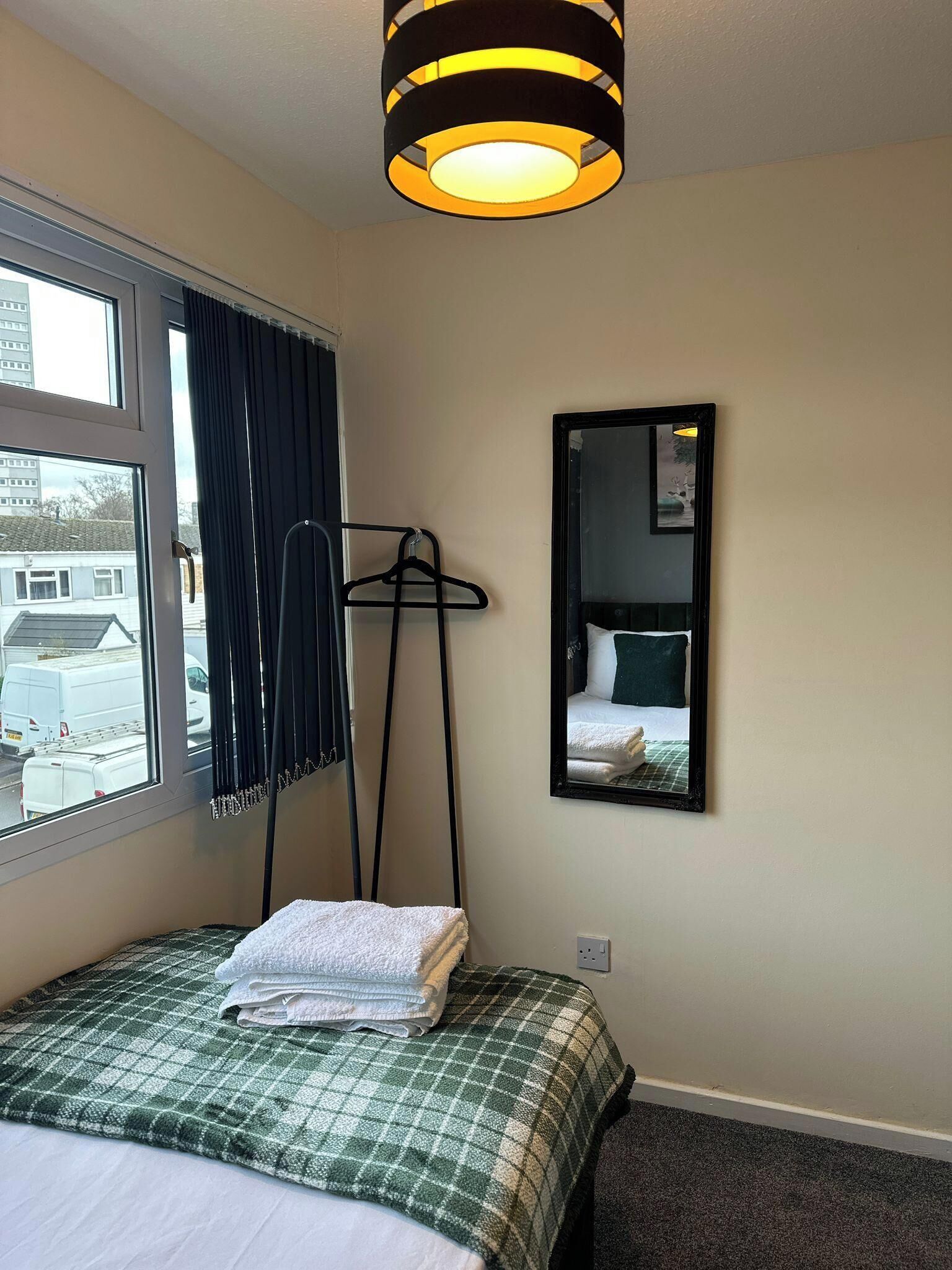 Quarto de hotel com cama, cabideiro, espelho e janela com cortinas.