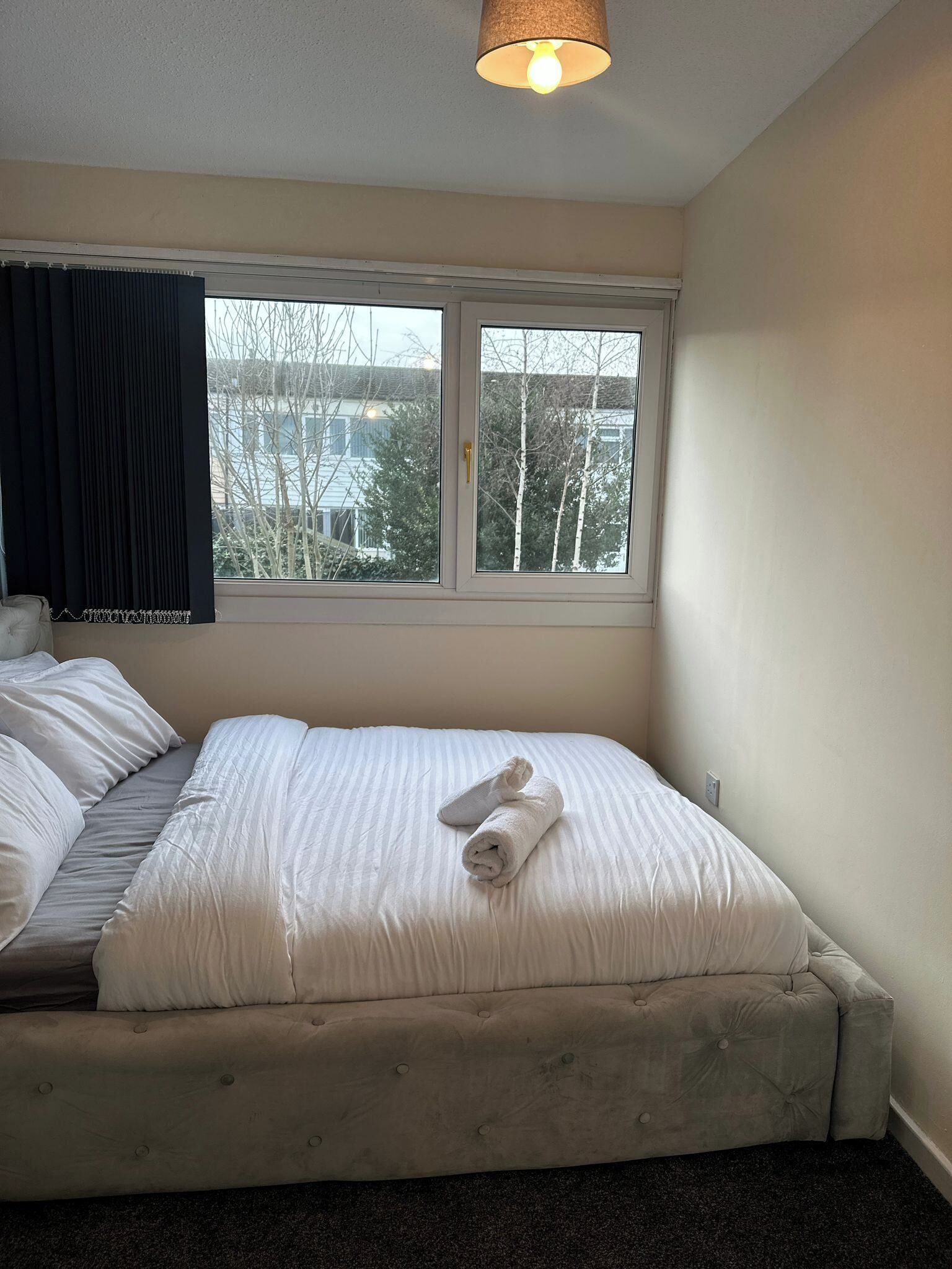 Cama bem arrumada com uma toalha enrolada, janela com cortinas e uma luminária no teto.