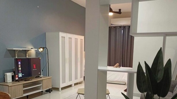 Uma sala de estar moderna com televisão de tela plana, um móvel de entretenimento de madeira, um armário branco, uma área de jantar com mesa e cadeiras, e uma grande planta em vaso.