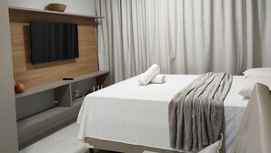 Quarto de hotel moderno com uma cama grande, uma televisão de tela plana na parede, uma mesinha de cabeceira e uma janela ampla com cortinas.