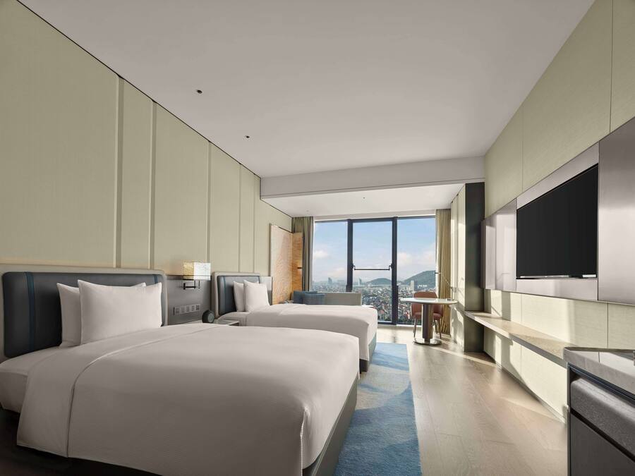 Quarto de hotel com duas camas, televisão, uma mesinha e vista para a cidade.