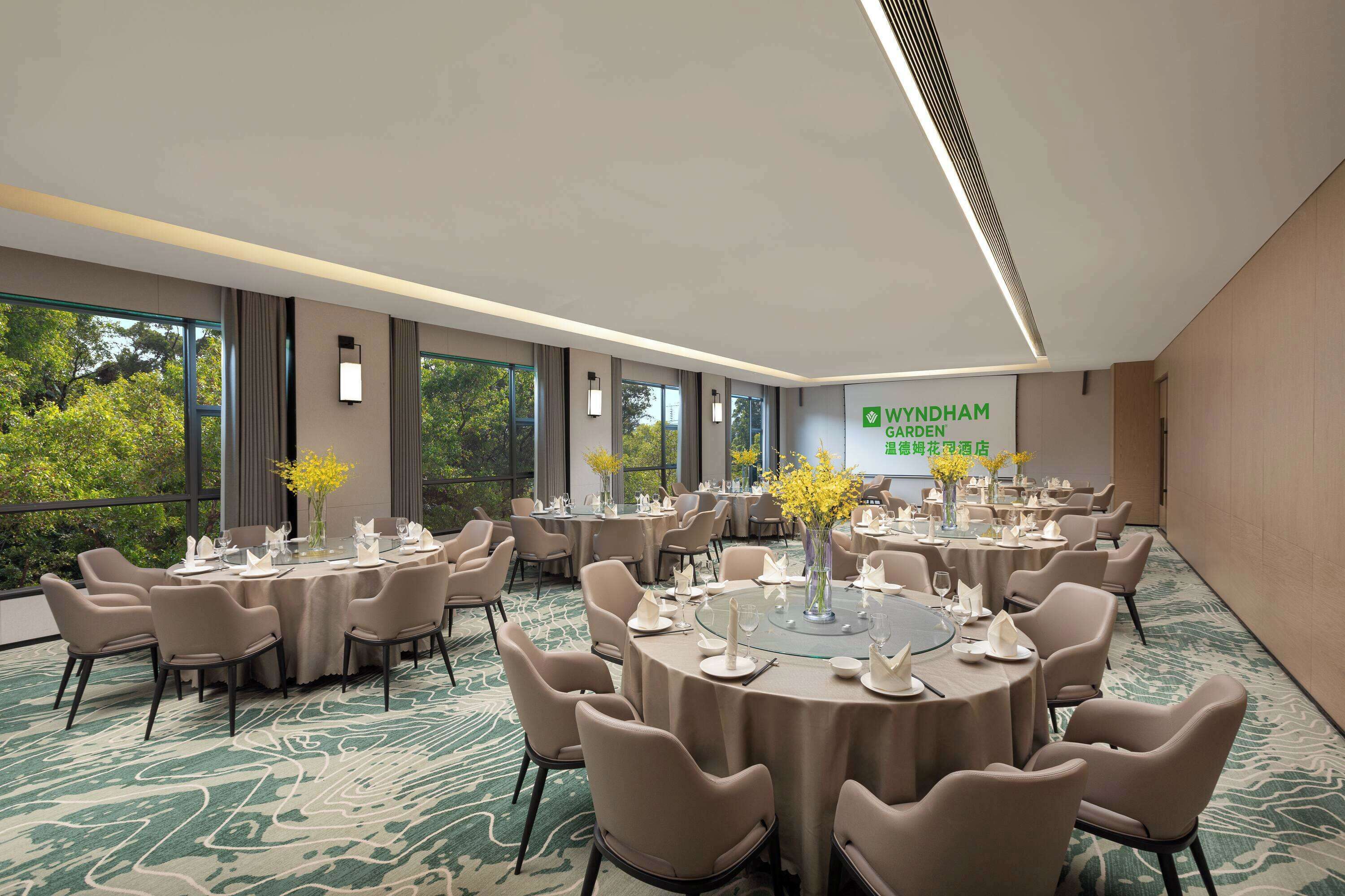 Sala de conferência com mesas redondas, cadeiras e uma tela de projetor exibindo 'WYNDHAM GARDEN'.