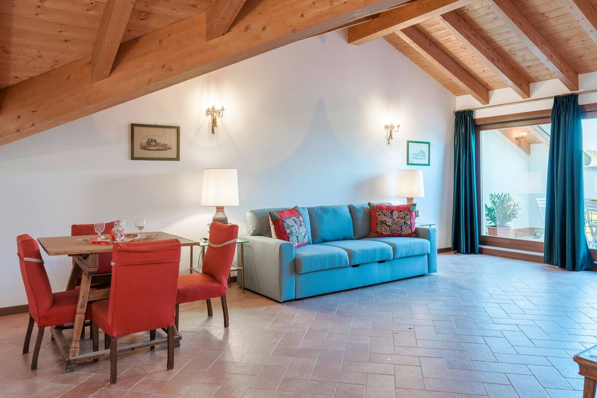 Uma sala de estar acolhedora com teto de madeira, um sofá azul, cadeiras vermelhas, uma mesa de jantar com copos e uma janela com cortinas.