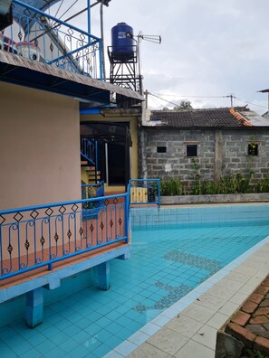 Uma piscina com deck em azulejos azuis, guarda-corpo de metal e um edifício de pedra ao fundo.