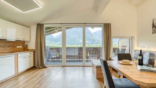 Uma cozinha moderna com bancada de madeira, área de refeições com mesa e cadeiras, e grandes portas de vidro deslizantes que dão para uma varanda com vista para a montanha.