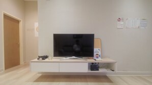 Uma sala de estar moderna com uma TV de tela plana em um móvel branco, uma pequena mesa com um vaso de vidro e uma porta à esquerda.