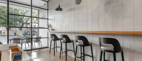 Uma área de bar moderna com balcão de madeira e banquetas pretas, grandes janelas e vista para a rua.