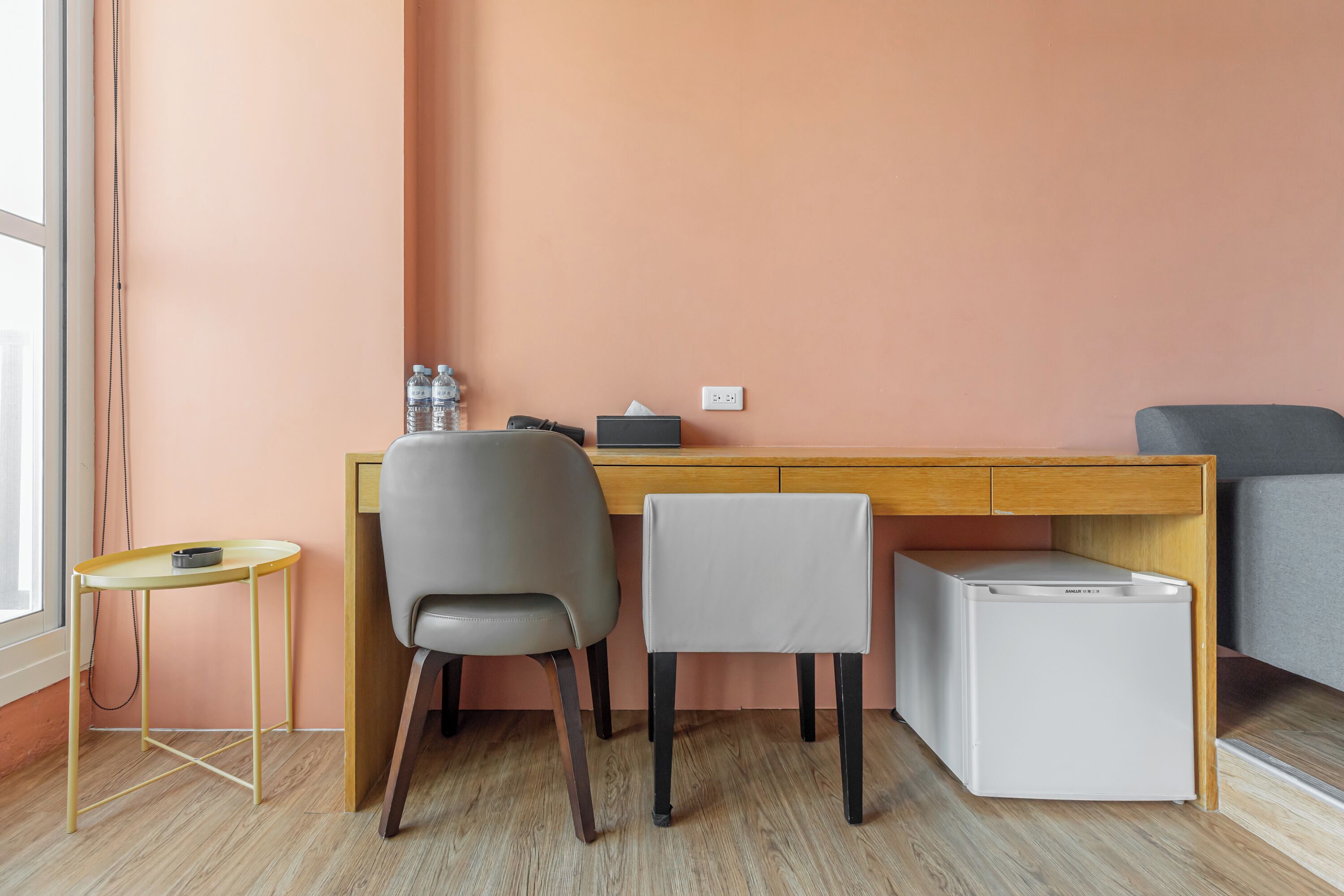 Quarto compacto com escrivaninha de madeira, duas cadeiras, uma mesinha e um mini-refrigerador.