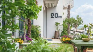 Um edifício com o número 20 grande na fachada, rodeado por vegetação exuberante e diversos vasos de plantas.