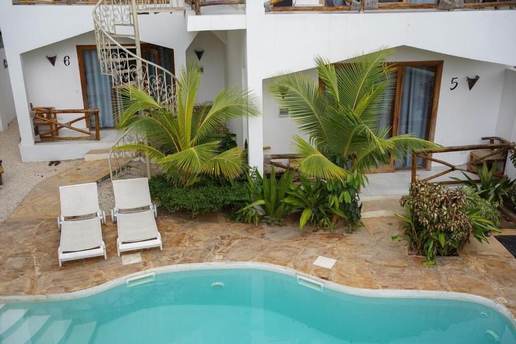 Área de piscina com espreguiçadeiras, palmeiras e uma escadaria que dá para uma varanda.
