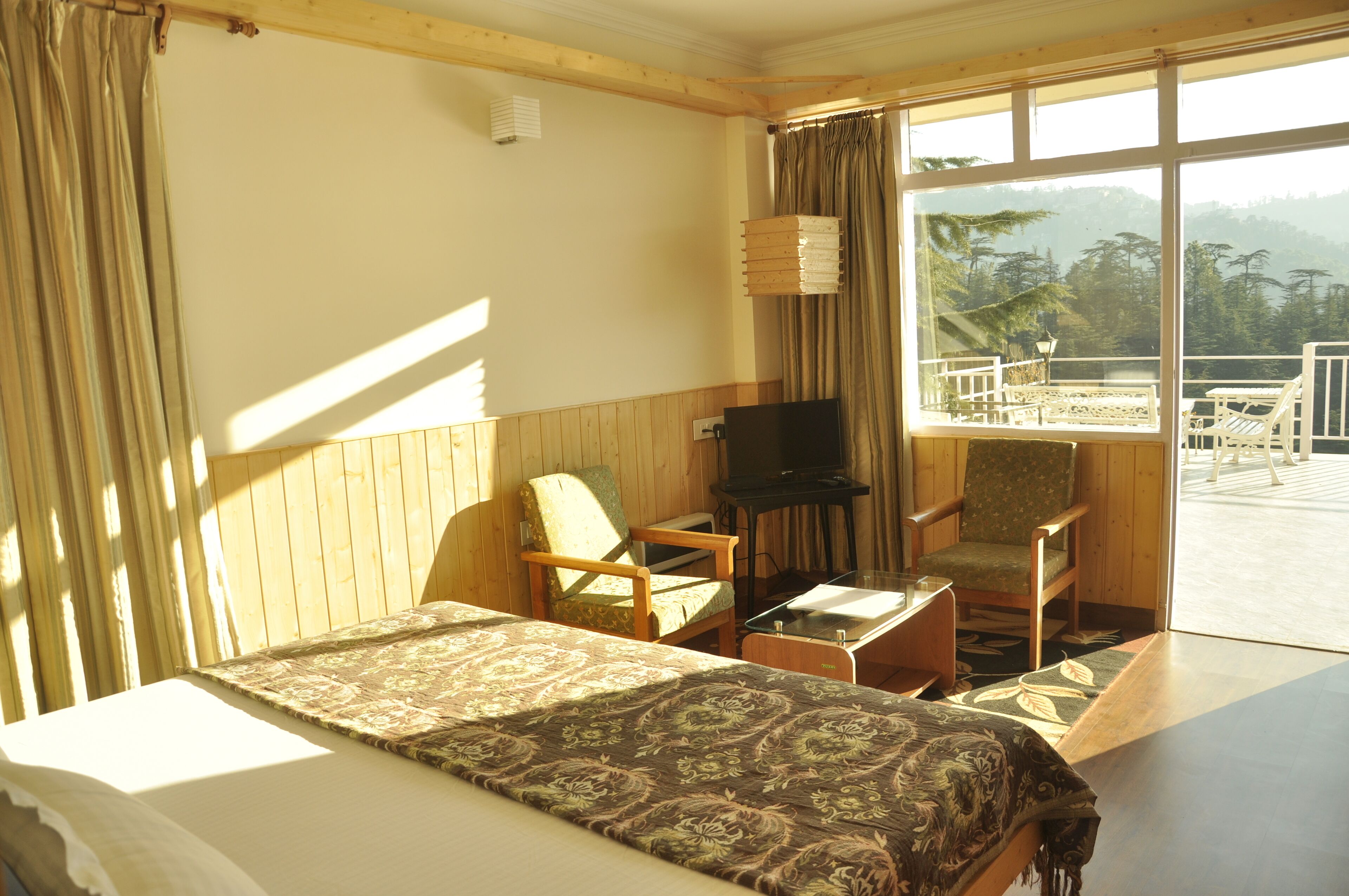 Quarto com cama, duas cadeiras, mesa pequena, televisão e uma janela grande com vista para uma varanda e árvores.