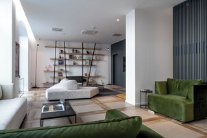 Uma sala de estar moderna com um sofá seccional, uma poltrona verde, um sofá branco e uma mesa de centro. Há uma estante com diversos objetos decorativos e um abajur.