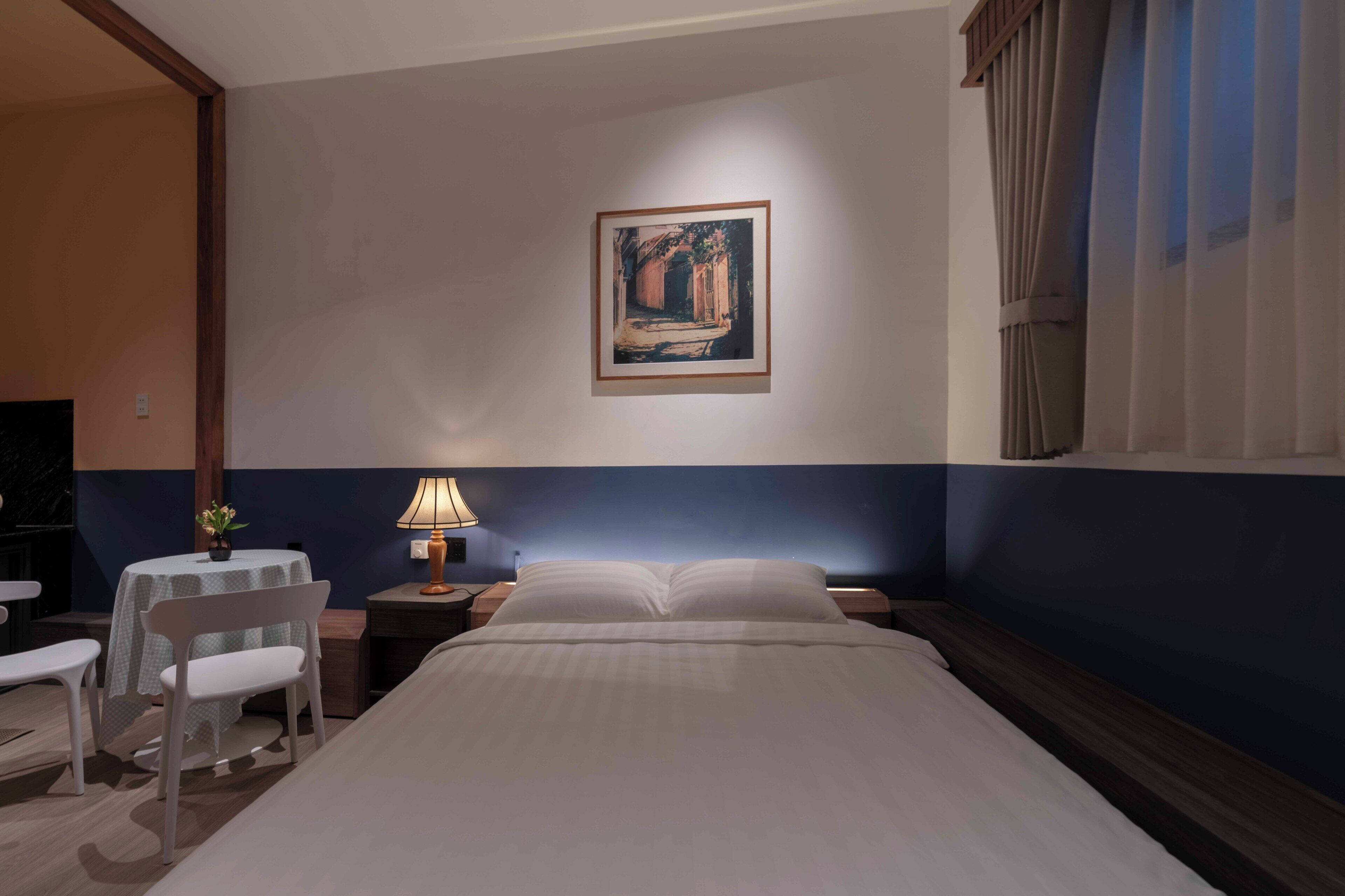 Quarto de hotel com cama, mesa de cabeceira com abajur, pequena mesa com cadeira e quadro na parede.