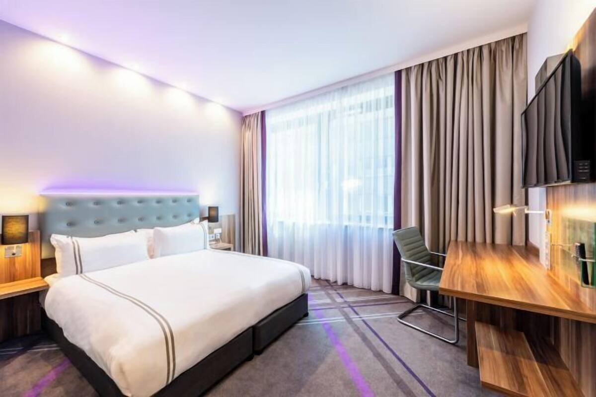 Quarto de hotel com cama, escrivaninha, cadeira, televisão e cortinas.