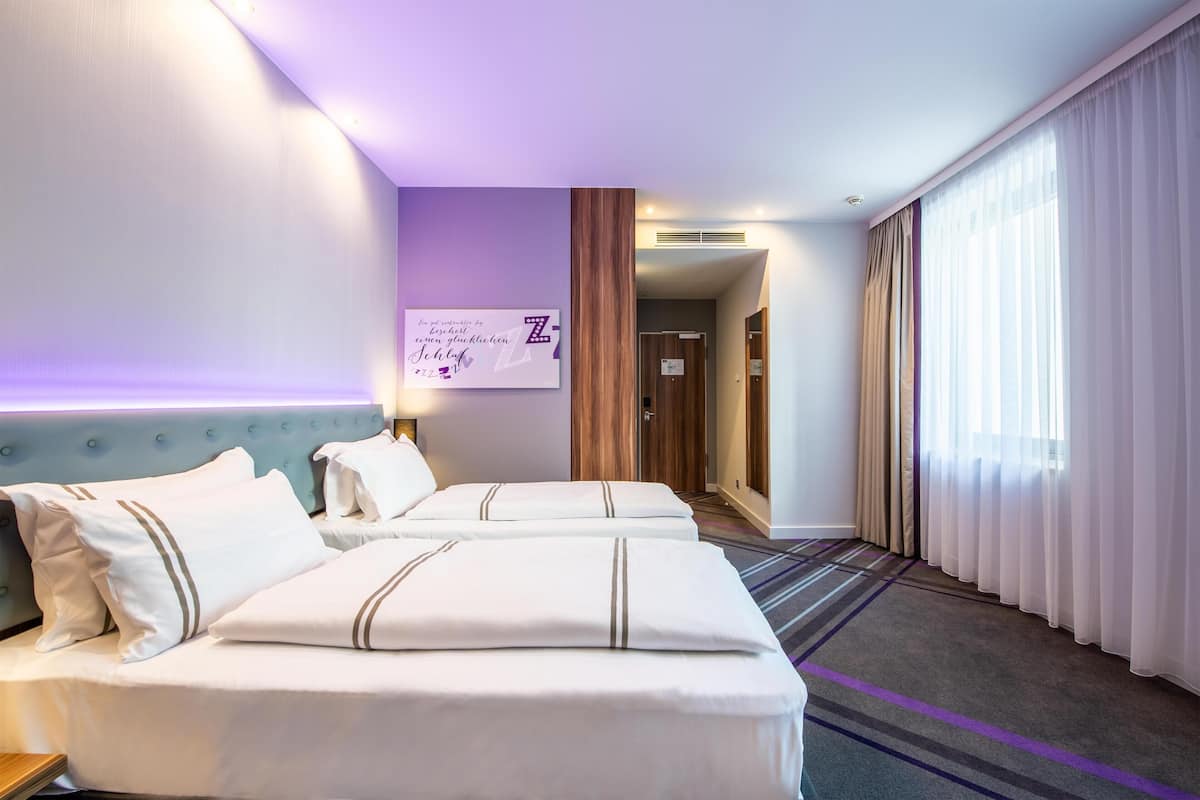 Quarto de hotel com duas camas, cabeceira, placa na parede e uma grande janela com cortinas.