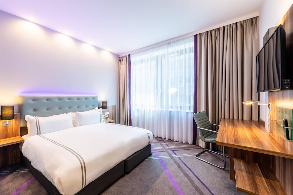 Quarto de hotel com cama, escrivaninha, cadeira, televisão e janela com cortinas.