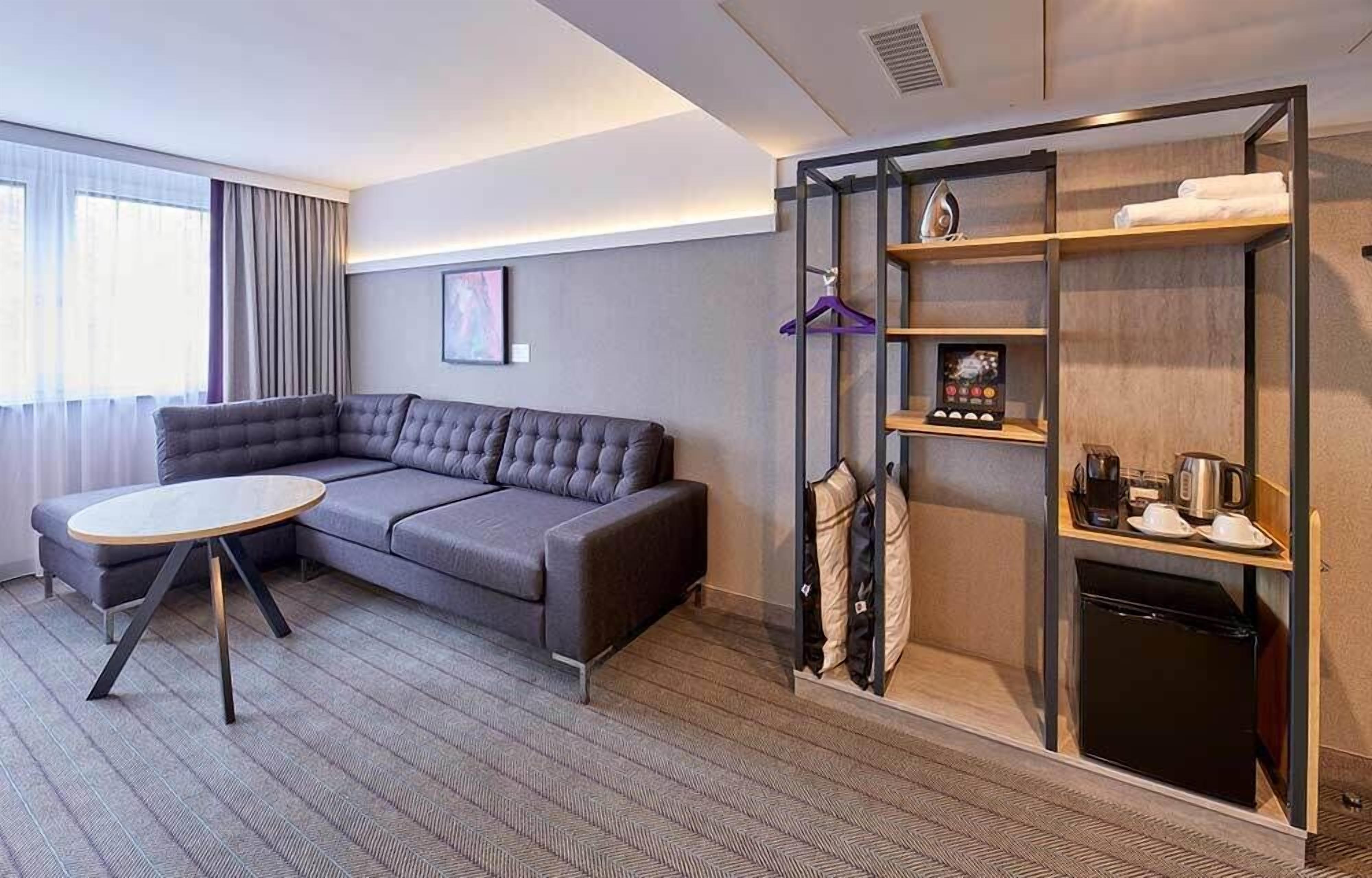 Quarto de hotel moderno com sofá modular cinza, mesa redonda, roupeiro embutido com prateleiras e um frigorífico preto pequeno.