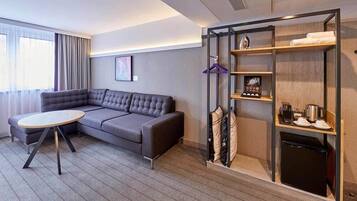 Quarto de hotel moderno com sofá modular cinza, mesa redonda, roupeiro embutido com prateleiras e um frigorífico preto pequeno.