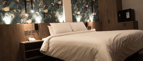 Quarto de hotel com uma cama grande, cabeceira de madeira e parede com papel de parede floral. Há uma mesinha de cabeceira com um telefone e um armário com uma TV.
