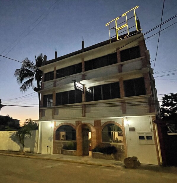 Um edifício de dois andares com uma placa indicando que é um hotel. Há várias janelas em cada andar, e o edifício tem um telhado plano com guarda-corpo no topo.