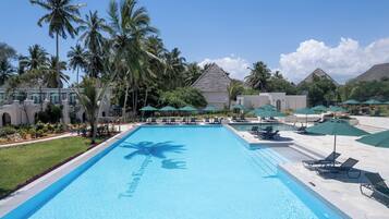 Piscina com espreguiçadeiras e guarda-sóis, cercada por palmeiras e edifícios com arquitetura tropical.