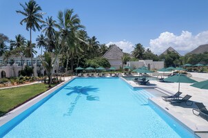 Piscina com espreguiçadeiras e guarda-sóis, cercada por palmeiras e edifícios com arquitetura tropical.
