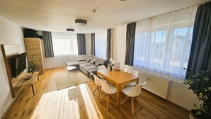 Uma sala de estar moderna com sofá, mesa de jantar e cadeiras. Há uma televisão em um suporte, uma planta e um radiador.