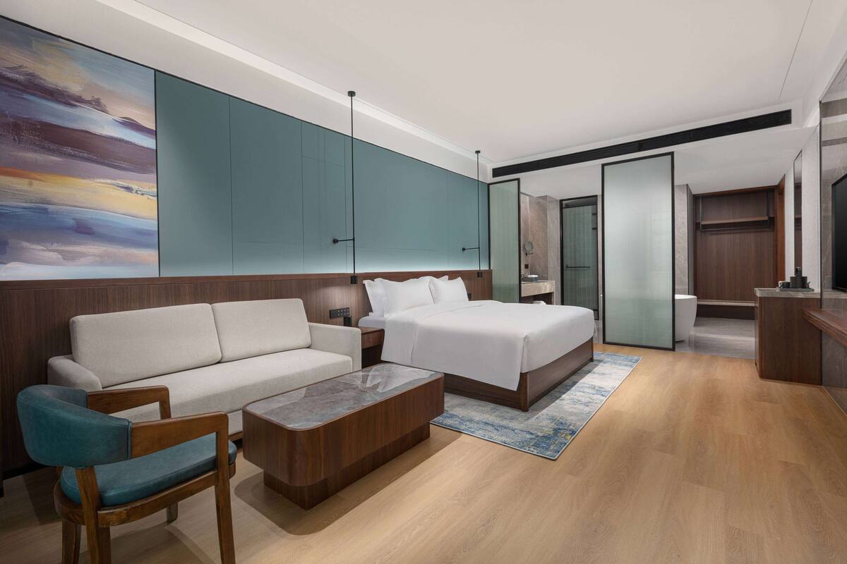 Quarto de hotel moderno com uma cama grande, um sofá, uma mesa de centro de madeira e uma grande pintura abstrata na parede.