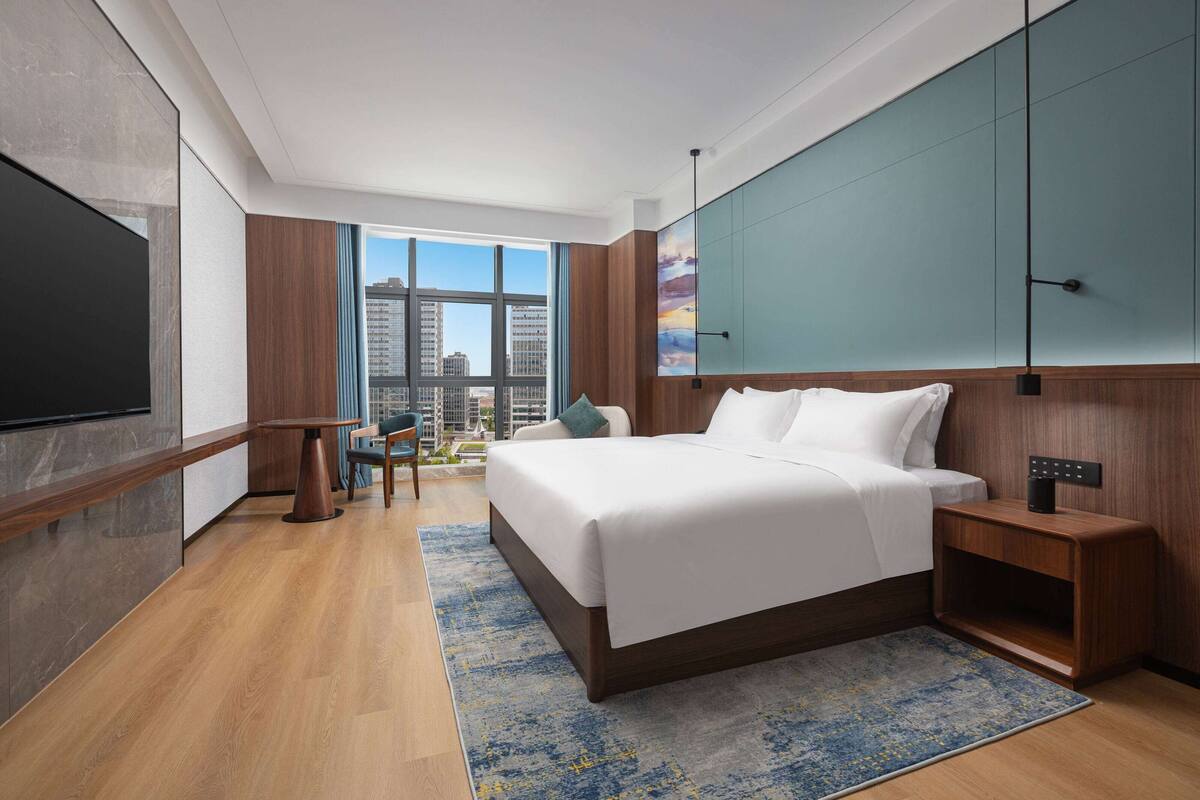 Quarto de hotel moderno com uma cama grande, televisão, escrivaninha e vista para a cidade.