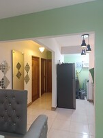 Um corredor com uma poltrona estofada cinza, piso de azulejos e uma geladeira ao fundo.