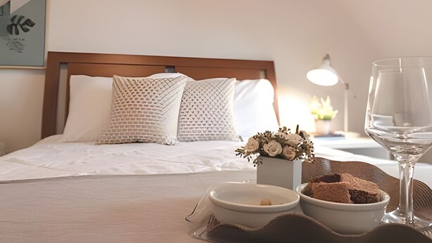 Cama bem arrumada com duas almofadas decorativas, um pequeno vaso com flores secas, uma tigela com sobremesa de chocolate e uma taça de vinho em uma mesa de cabeceira.