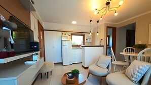 Uma sala de estar moderna com um sofá, poltronas, uma mesa de centro e uma televisão. Ao fundo, há uma área de cozinha com uma geladeira e armários.