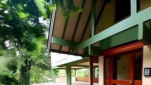 Uma área externa coberta com chão de azulejos, uma porta vermelha e fachada de prédio verde.