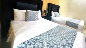 Quarto de hotel com duas camas, cada uma com colcha e almofadas estampadas. Há uma mesinha de cabeceira com abajur entre as camas.