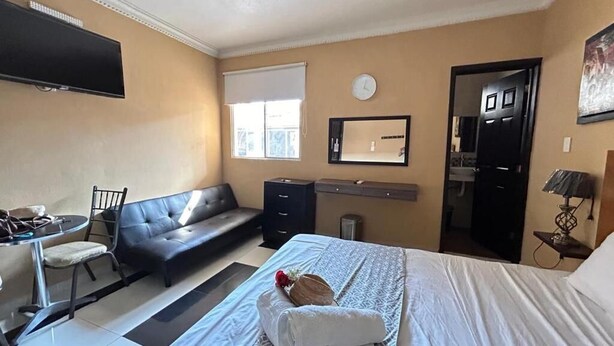 Quarto de hotel com cama, sofá, escrivaninha, televisão, relógio e espelho.