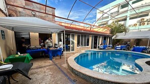 Uma área de piscina ao ar livre com um espaço de estar coberto, mesas e um edifício ao fundo.