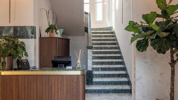 Uma área de recepção com balcão de madeira, um telefone e uma planta em vaso. Escada com piso de mármore leva a um nível superior.