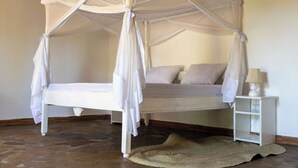 Uma cama de dossel branca com mesa de cabeceira e abajur, tapete tecido e chão de pedra.