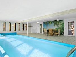 Uma piscina com deck azulejado, área de refeições com cadeiras de vime e um mural com tema tropical.