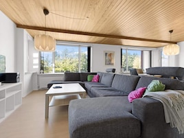 Uma sala de estar moderna com um amplo sofá seccional cinza, uma mesa de centro branca e teto de madeira. Janelas grandes oferecem luz natural e vista para o verde lá fora.