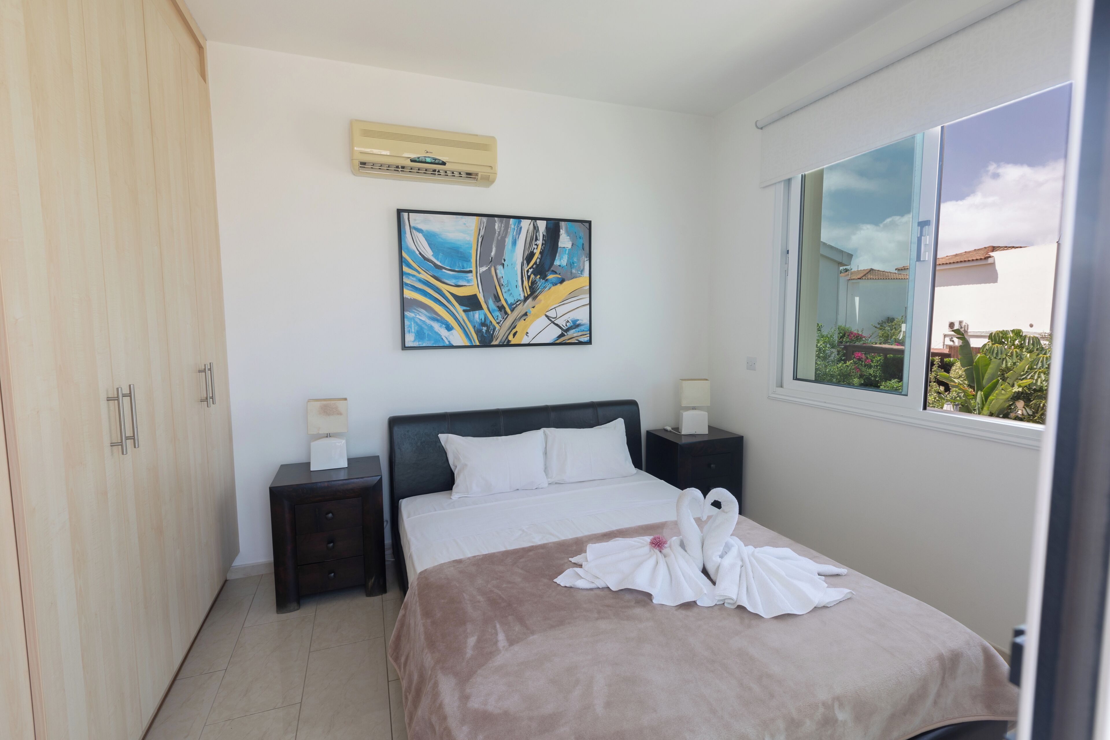 Um quarto com cama, mesa de cabeceira, guarda-roupa, pintura e janela com vista.
