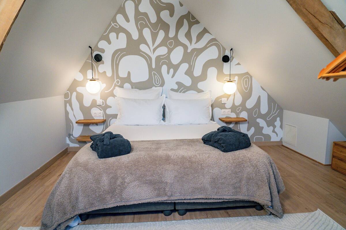 Quarto com teto inclinado, cama com duas almofadas, cobertor e duas mesas de cabeceira com abajures.