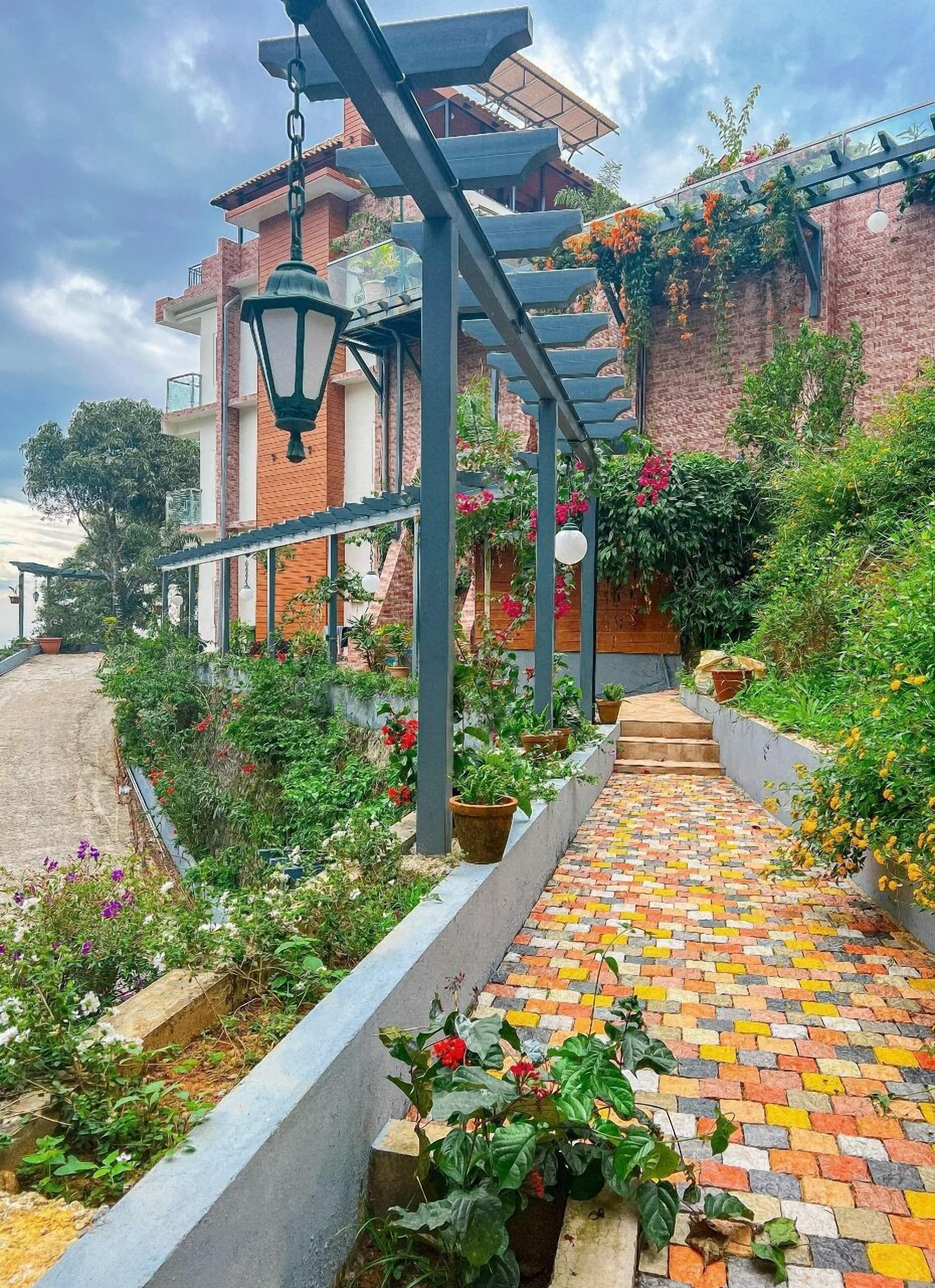 Um caminho no jardim leva a um edifício com fachada de tijolos, rodeado por diversas plantas e flores.