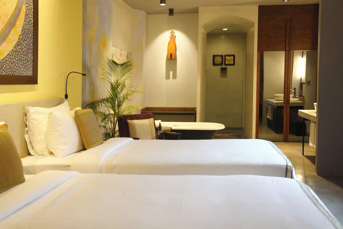 Quarto de hotel com duas camas, área de estar com mesa e cadeiras, banheiro pequeno e vista para o corredor.
