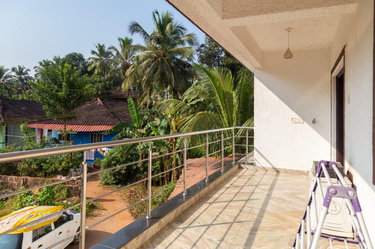 Varanda com guarda-corpo de metal, soalho em azulejos e vista para uma área residencial com casas e palmeiras.