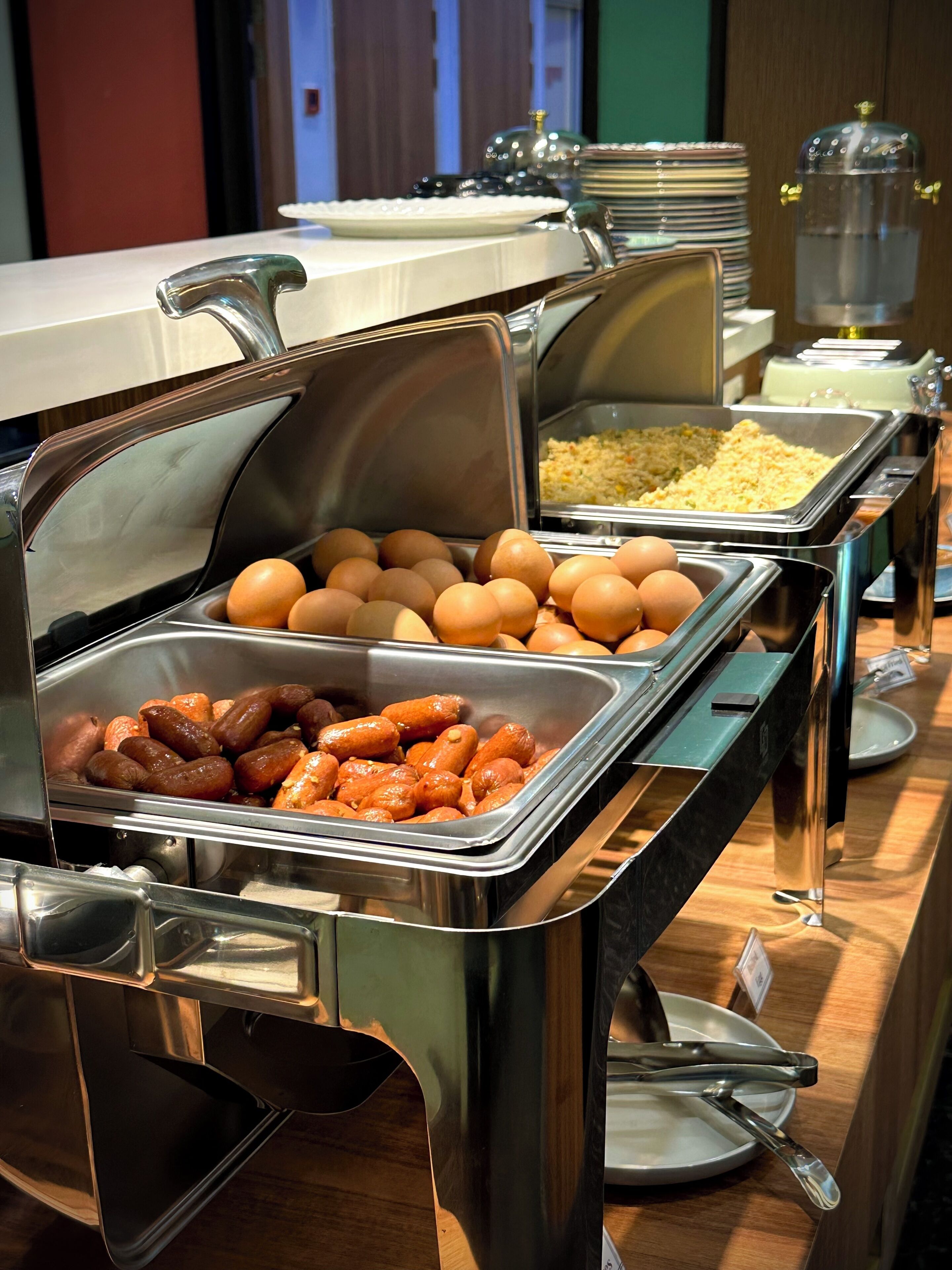 Buffet com diversas opções de comida, incluindo ovos, linguiça e pratos de arroz.